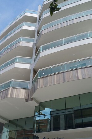 Um edifício de vários andares com design moderno, com varandas e guarda-corpos de vidro, e um logotipo visível na parte inferior da fachada.