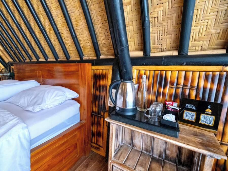 Quarto com cama, cabeceira de madeira, teto de bambu e uma pequena mesa com chaleira e xícara.