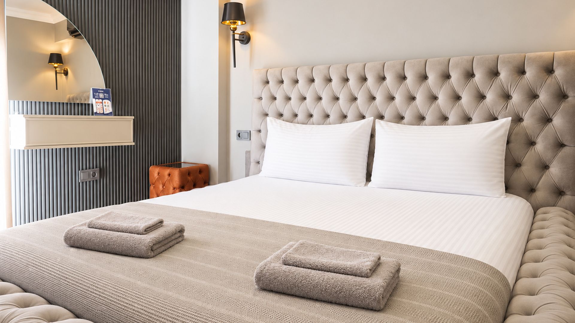 Quarto de hotel com uma cama grande, duas almofadas, cabeceira, mesa de cabeceira, abajur e uma toalha dobrada na cama.