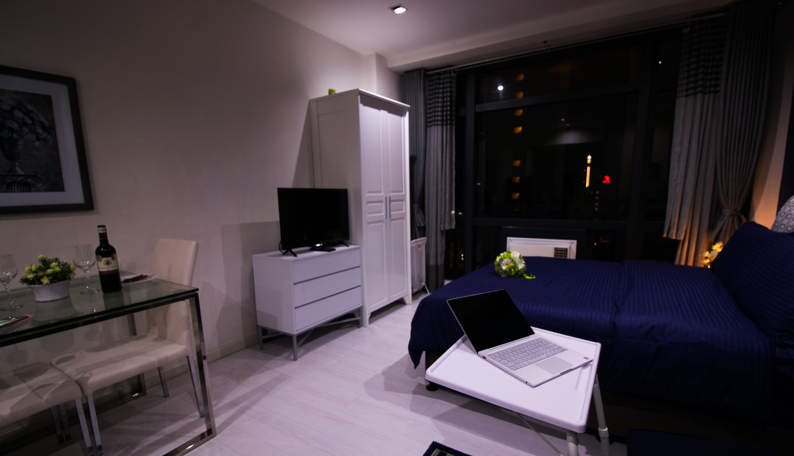 Quarto de hotel moderno com escrivaninha de vidro, um laptop sobre uma mesinha, cama com cobertura azul escura, guarda-roupa branco e uma janela grande com cortinas.