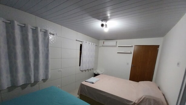Quarto pequeno com uma cama de solteiro, colcha azul, cortinas brancas, porta de madeira e luminária no teto.
