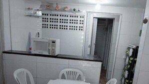 Uma cozinha compacta com armários brancos, micro-ondas e uma pequena área de refeições com cadeiras de plástico.