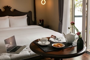 Uma cama bem arrumada com um laptop, uma chávena de café, um bule, um prato de pastelaria e um vaso com flores numa mesa de cabeceira.