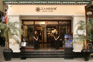 Entrada do Hotel Lamejor Luxury, com um letreiro de restaurante, dois funcionários e plantas em vasos.