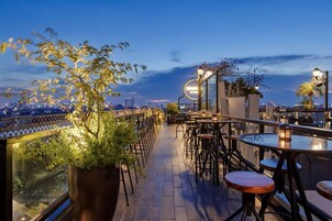 Um bar no terraço com mesas ao ar livre, vista da cidade e iluminação decorativa.