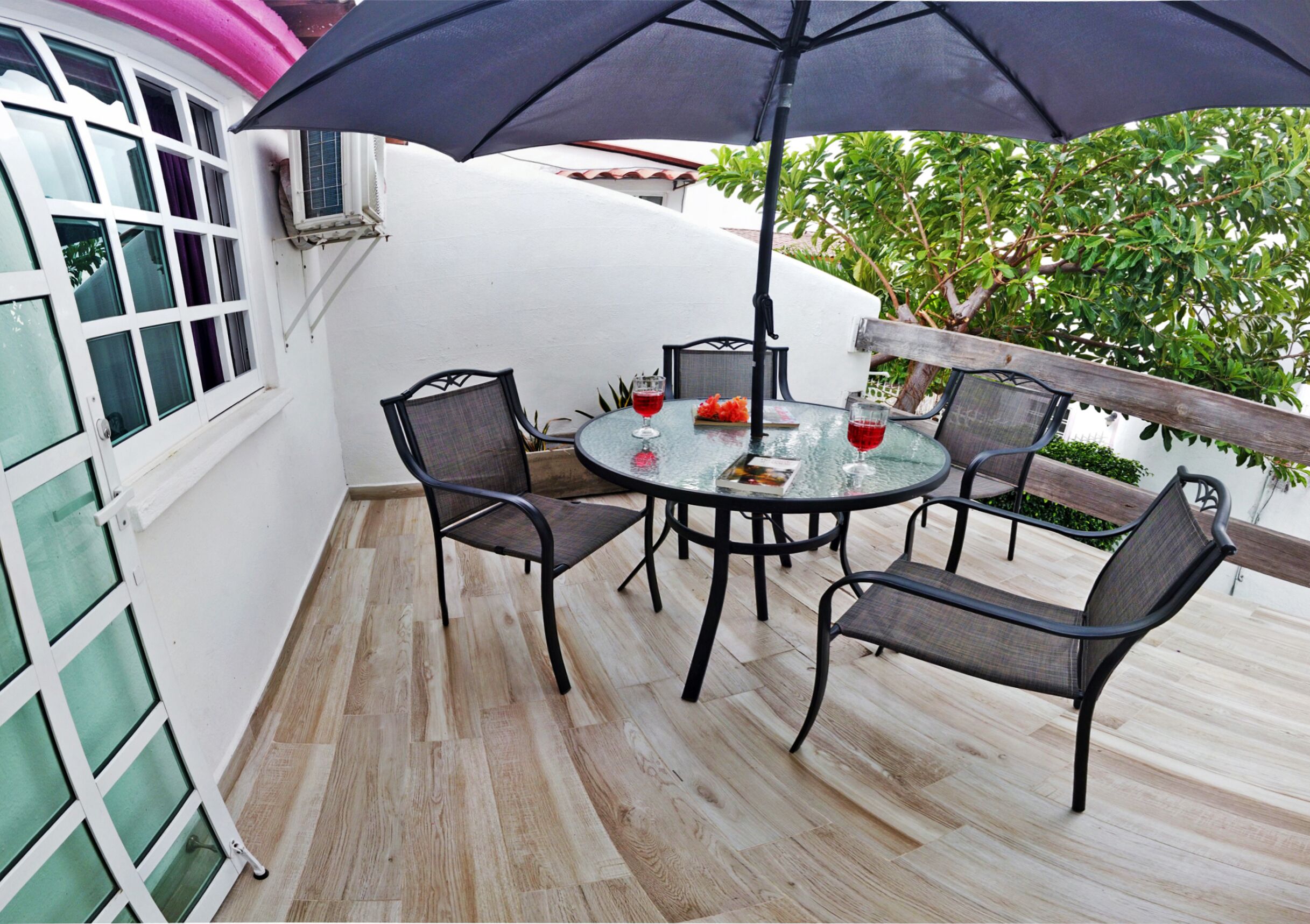 Mesa redonda com quatro cadeiras e um guarda-sol grande, cercada por um deck de madeira com uma parede branca e uma janela com painéis de vidro verde.