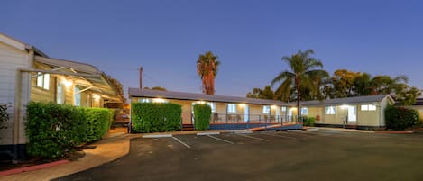 Um motel com vários quartos, área de estacionamento coberta e um pequeno jardim com arbustos e uma palmeira.