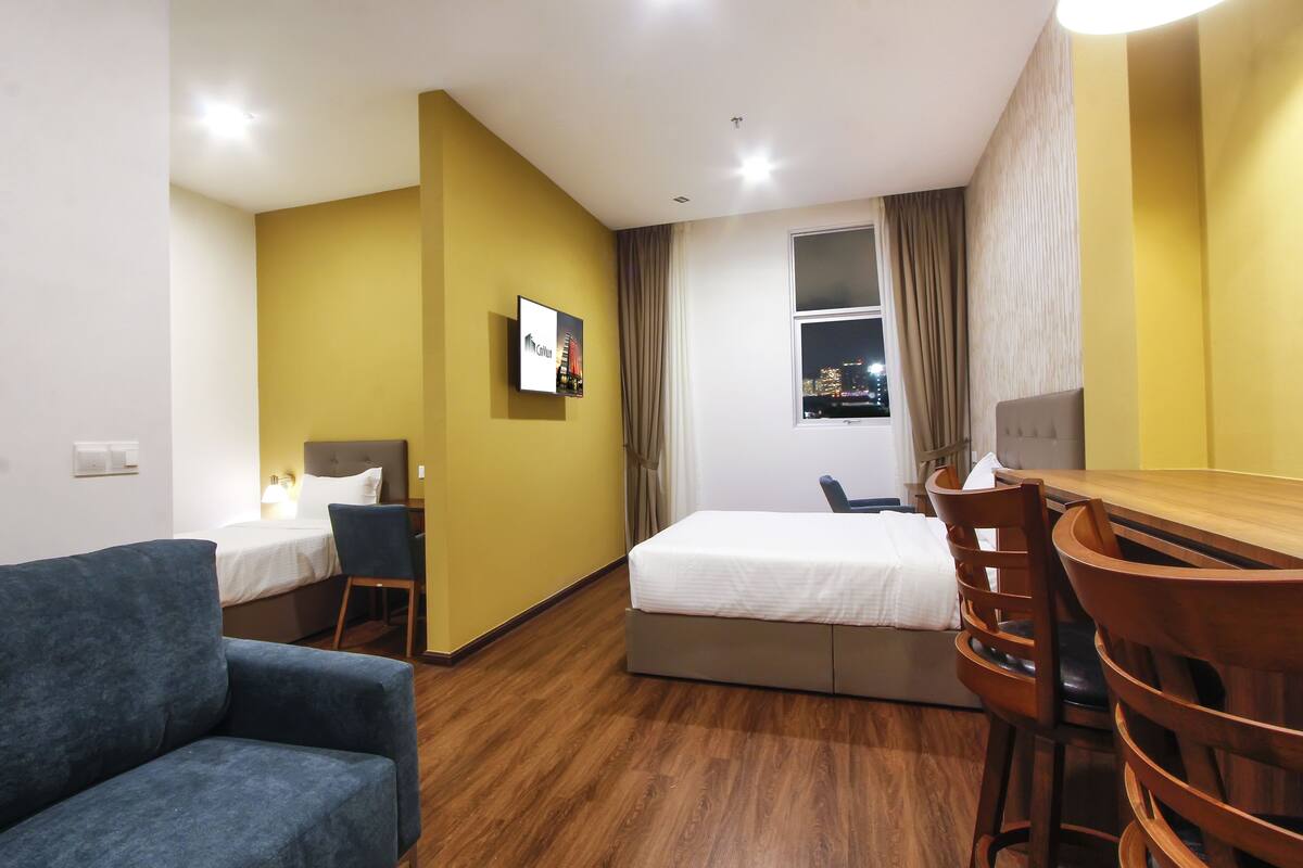 Quarto de hotel com cama, escrivaninha, cadeira, sofá e televisão na parede.