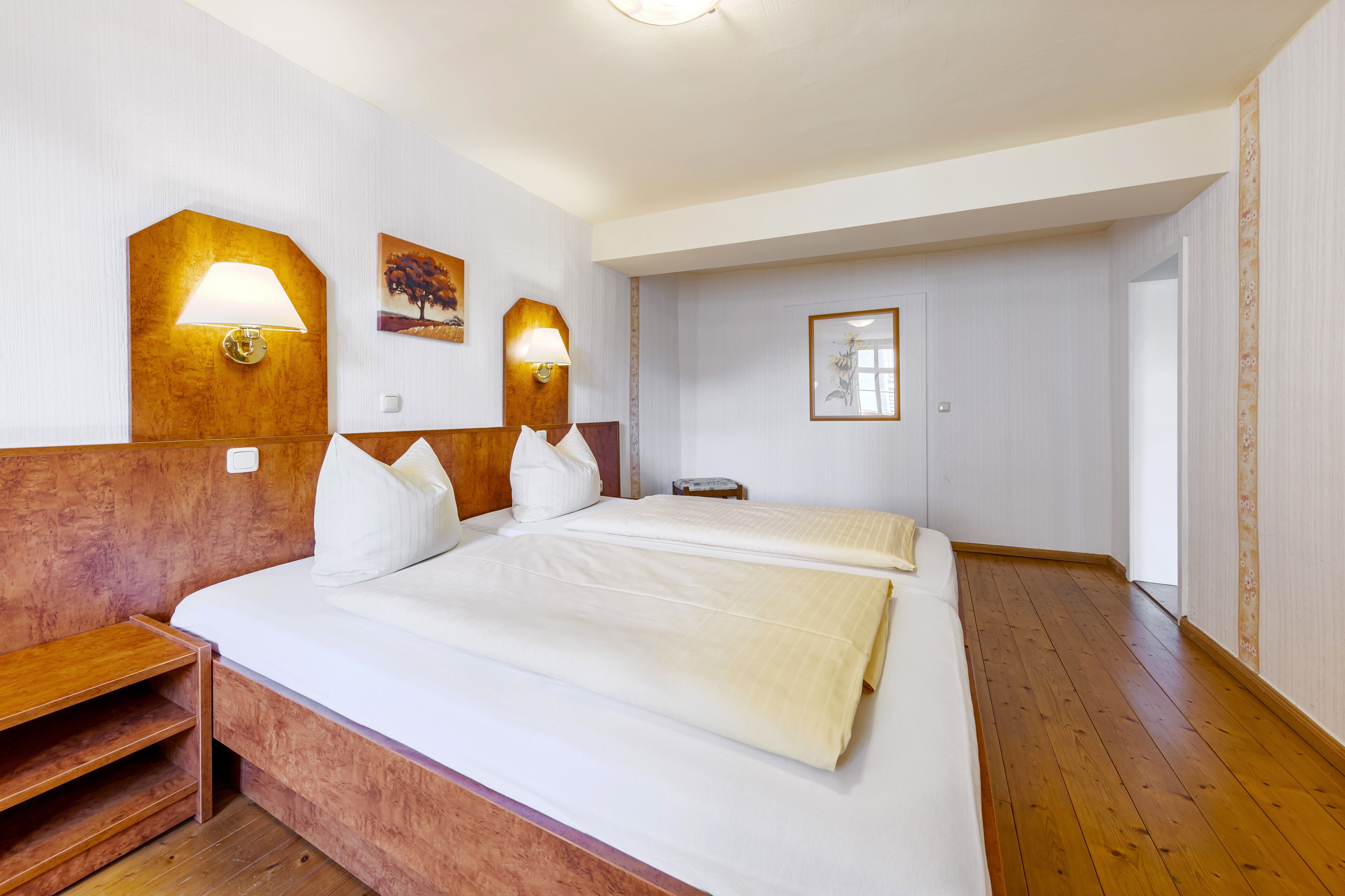 Quarto de hotel com duas camas, cabeceiras de madeira, uma mesinha de cabeceira e luminárias fixadas na parede.