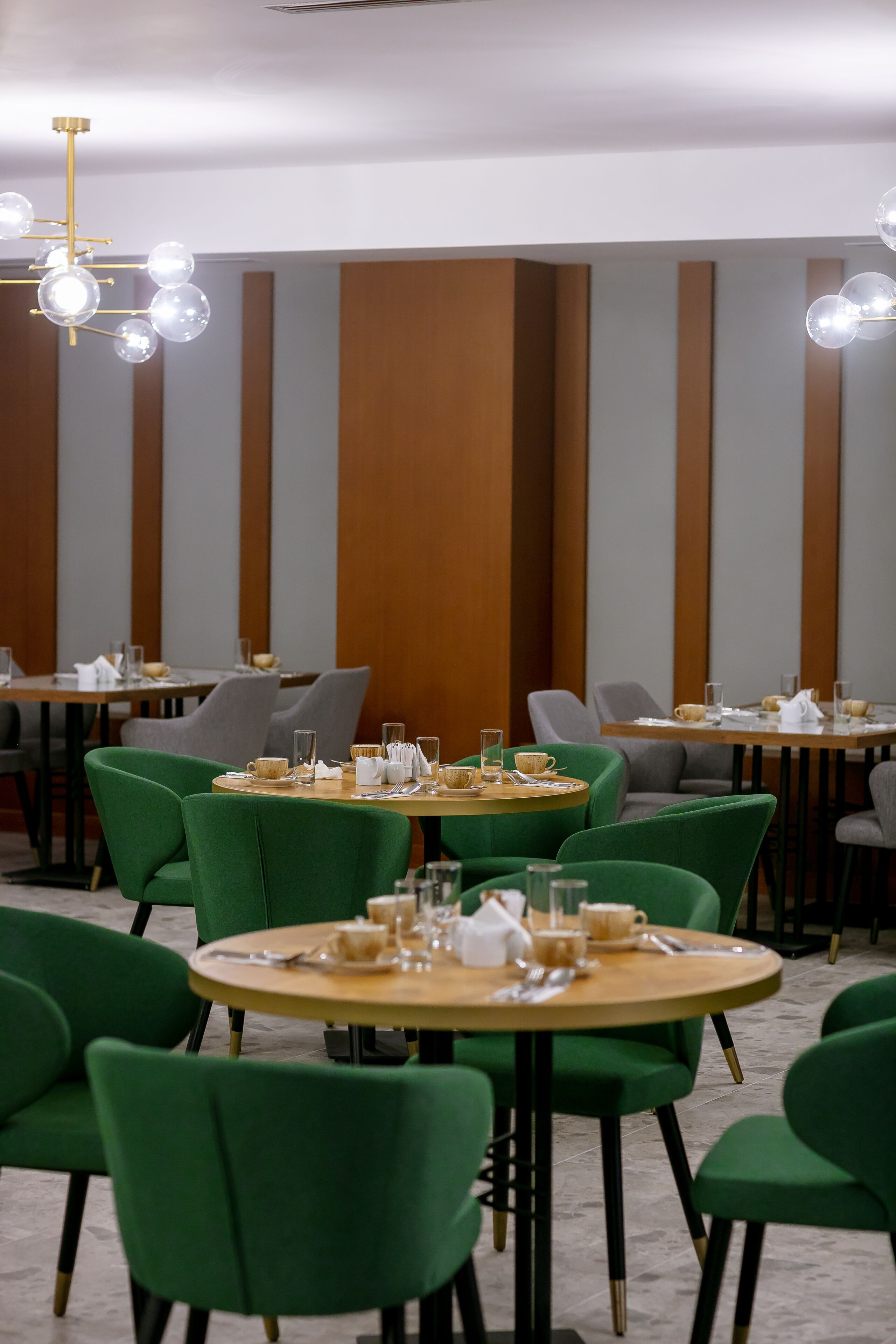 Uma sala de jantar com mesas redondas, cadeiras estofadas em verde e um lustre moderno.