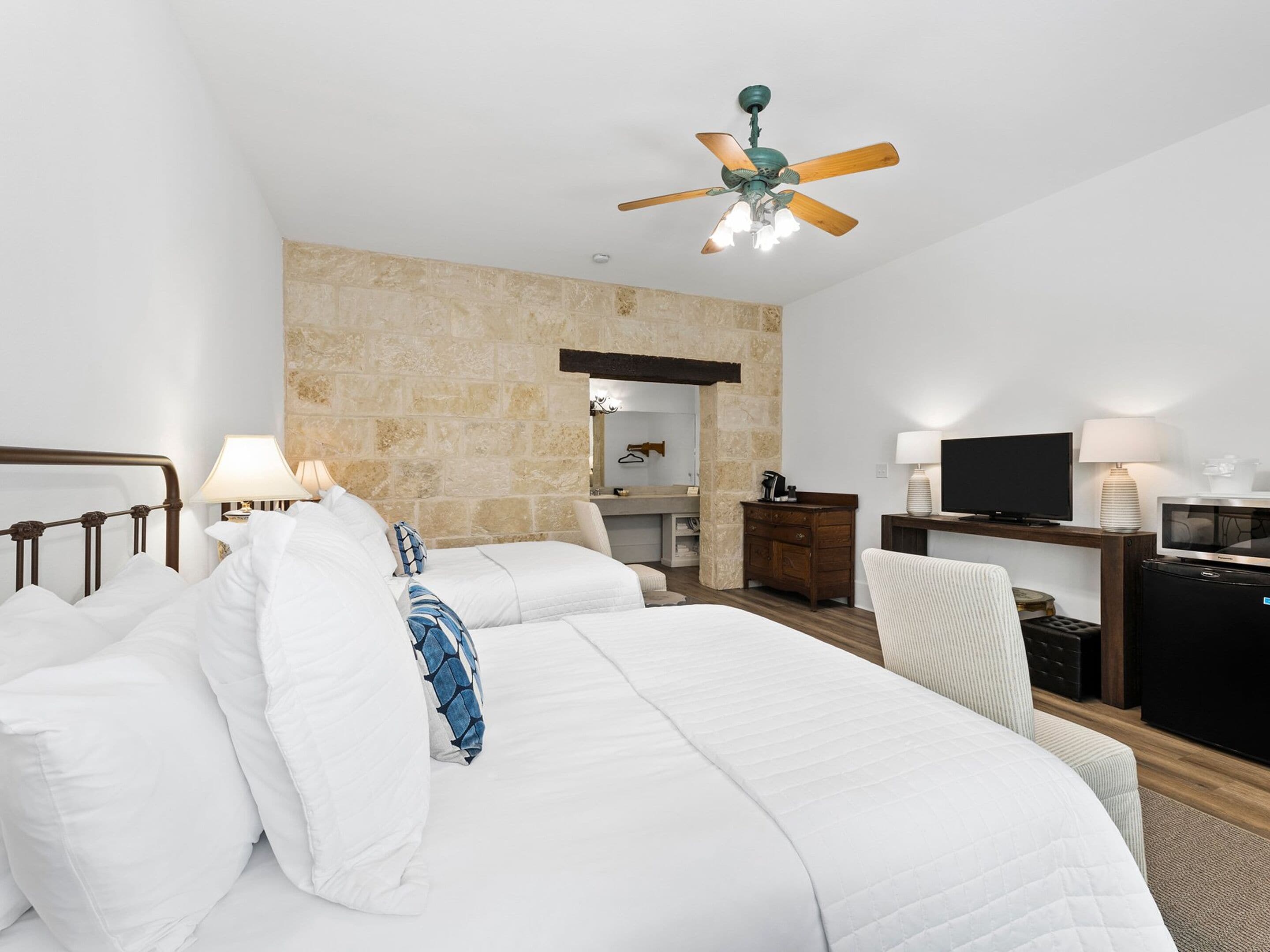 Quarto de hotel com uma cama grande, uma escrivaninha com televisão, ventoinha no teto e parede de detalhes em pedra.
