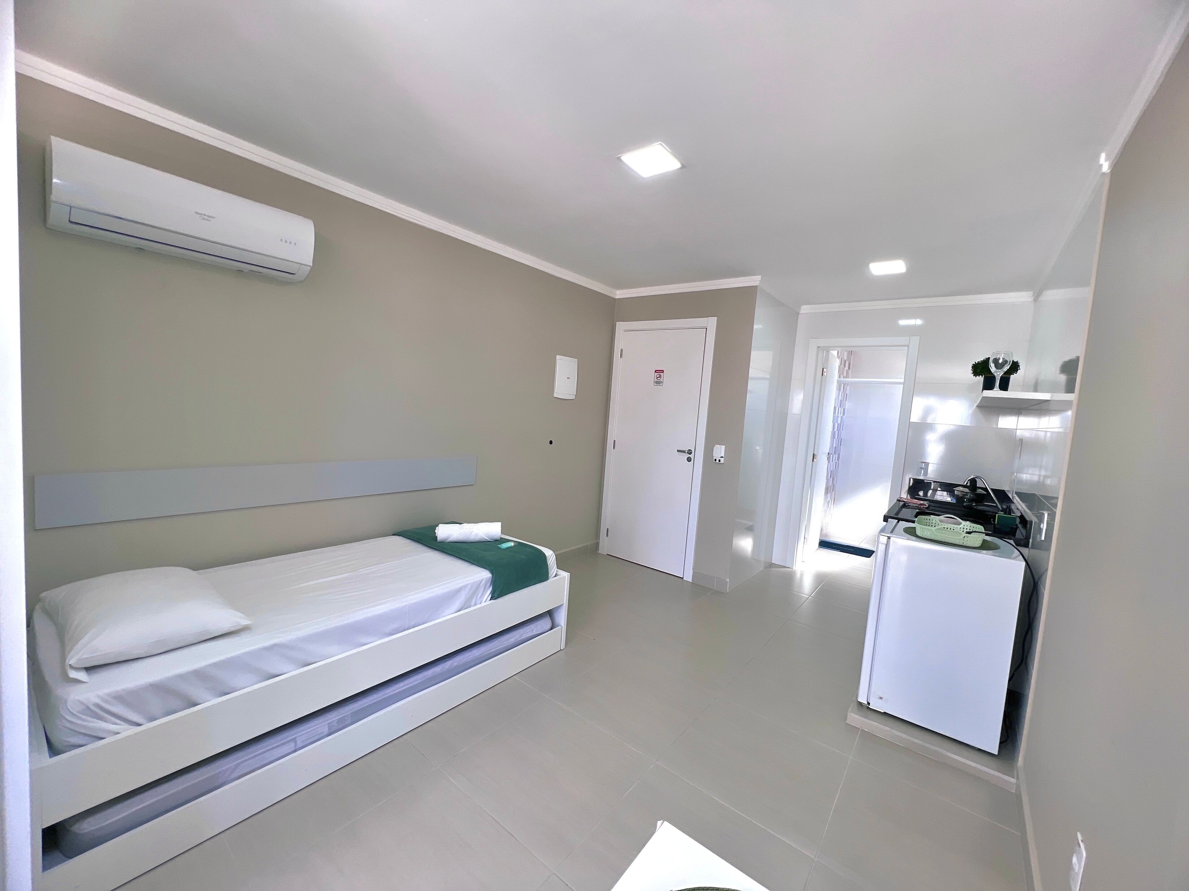 Quarto de hotel com uma cama de solteiro, uma kitchenette pequena e uma porta que dá para outro quarto.