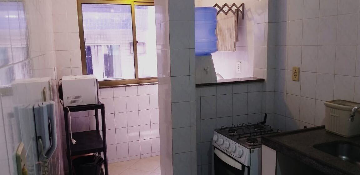 Uma cozinha pequena com paredes em azulejos brancos, fogão a gás, pia e janela com cortinas.