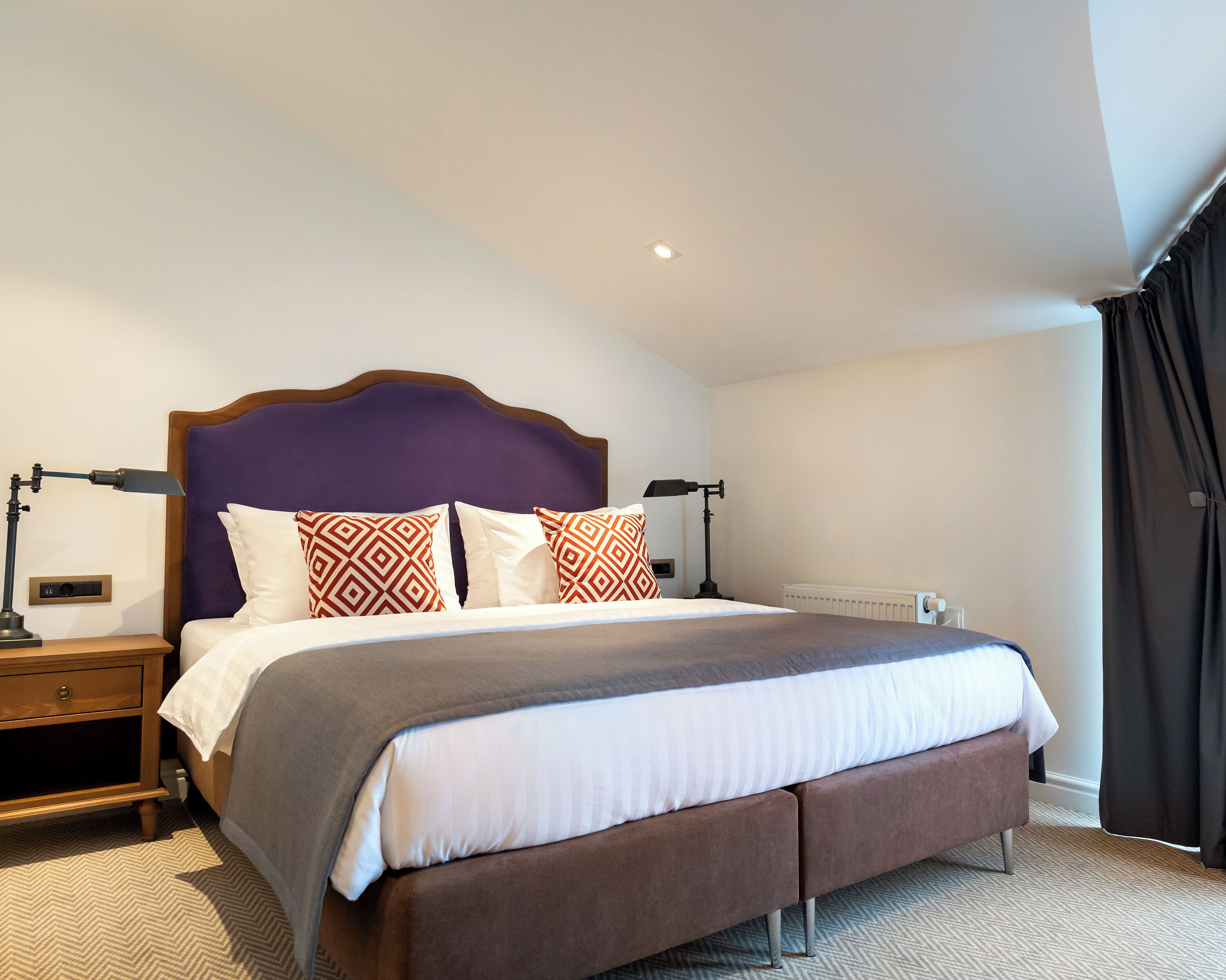 Quarto de hotel moderno com uma cama grande, duas mesas de cabeceira e uma janela com cortinas.