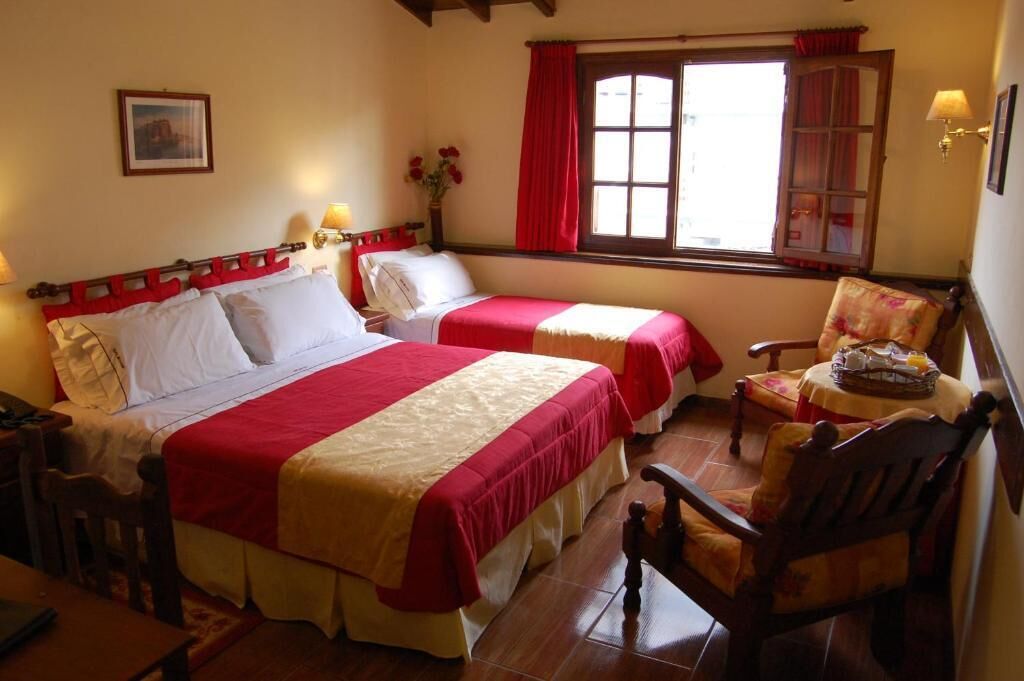 Quarto de hotel com duas camas, uma cadeira de madeira, uma mesa com uma cesta e uma janela com cortinas vermelhas.