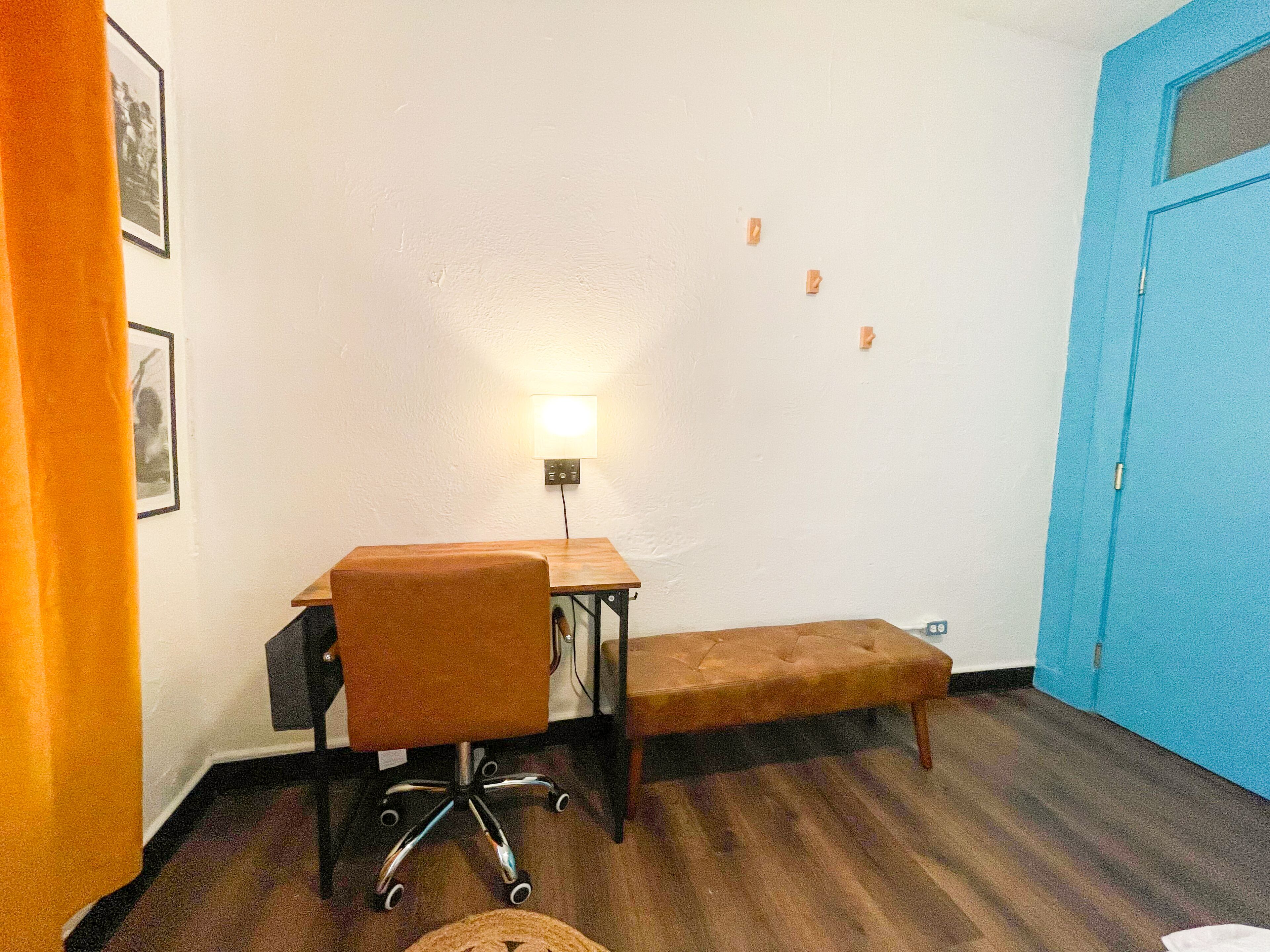 Quarto pequeno com escrivaninha, cadeira e banco. Há uma luminária na parede, uma porta e um quadro.