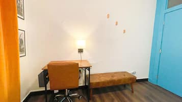 Quarto pequeno com escrivaninha, cadeira e banco. Há uma luminária na parede, uma porta e um quadro.