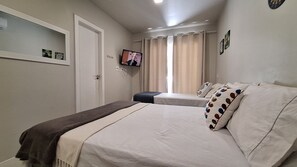 Quarto com duas camas, televisão na parede e janela com cortinas.