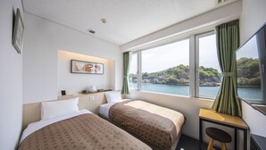 Quarto de hotel com duas camas, uma escrivaninha e janela com vista para a água e vegetação.