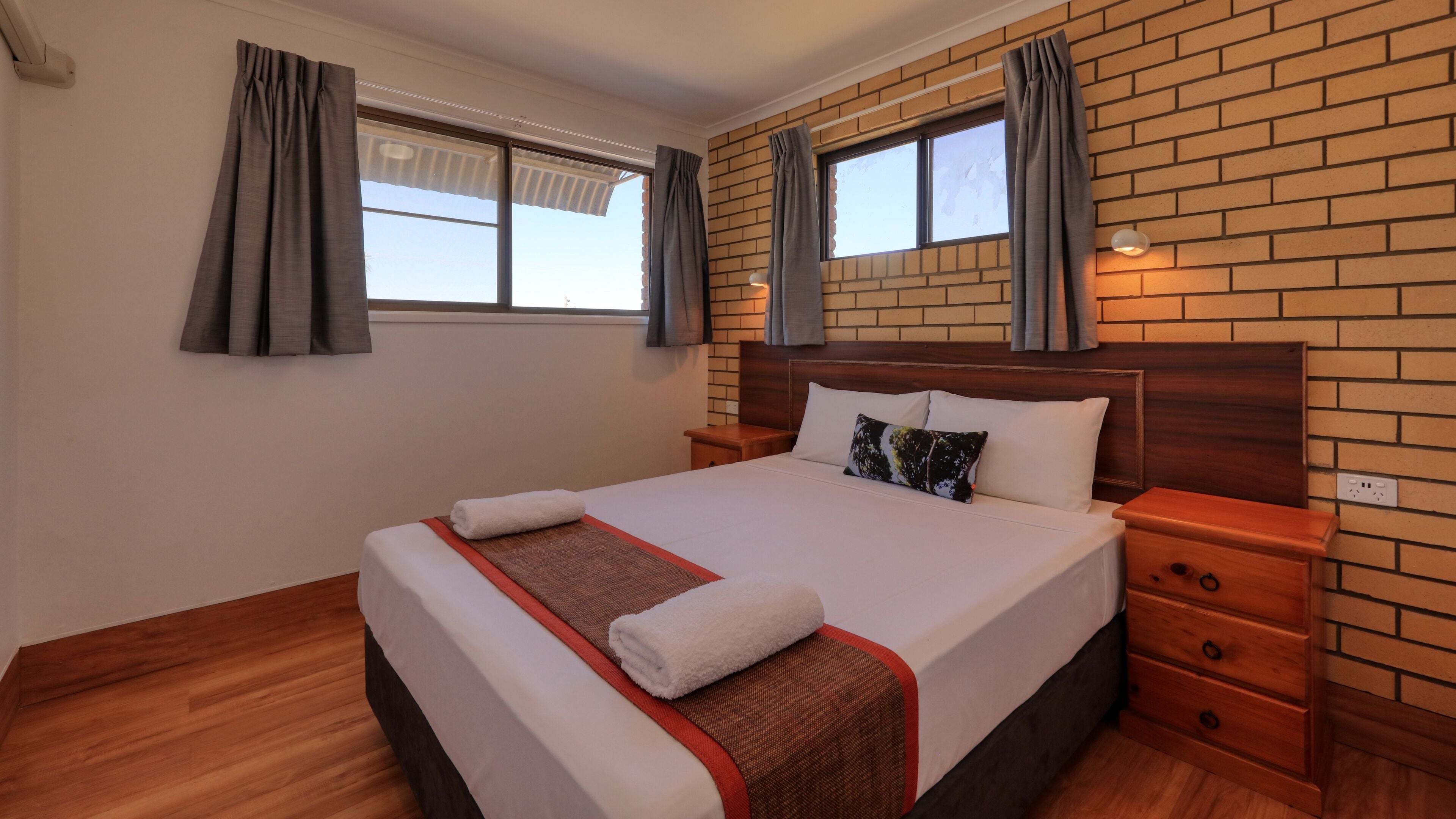 Quarto de hotel com uma cama grande, cabeceira de madeira, duas mesas de cabeceira e uma parede com padrão de tijolos.