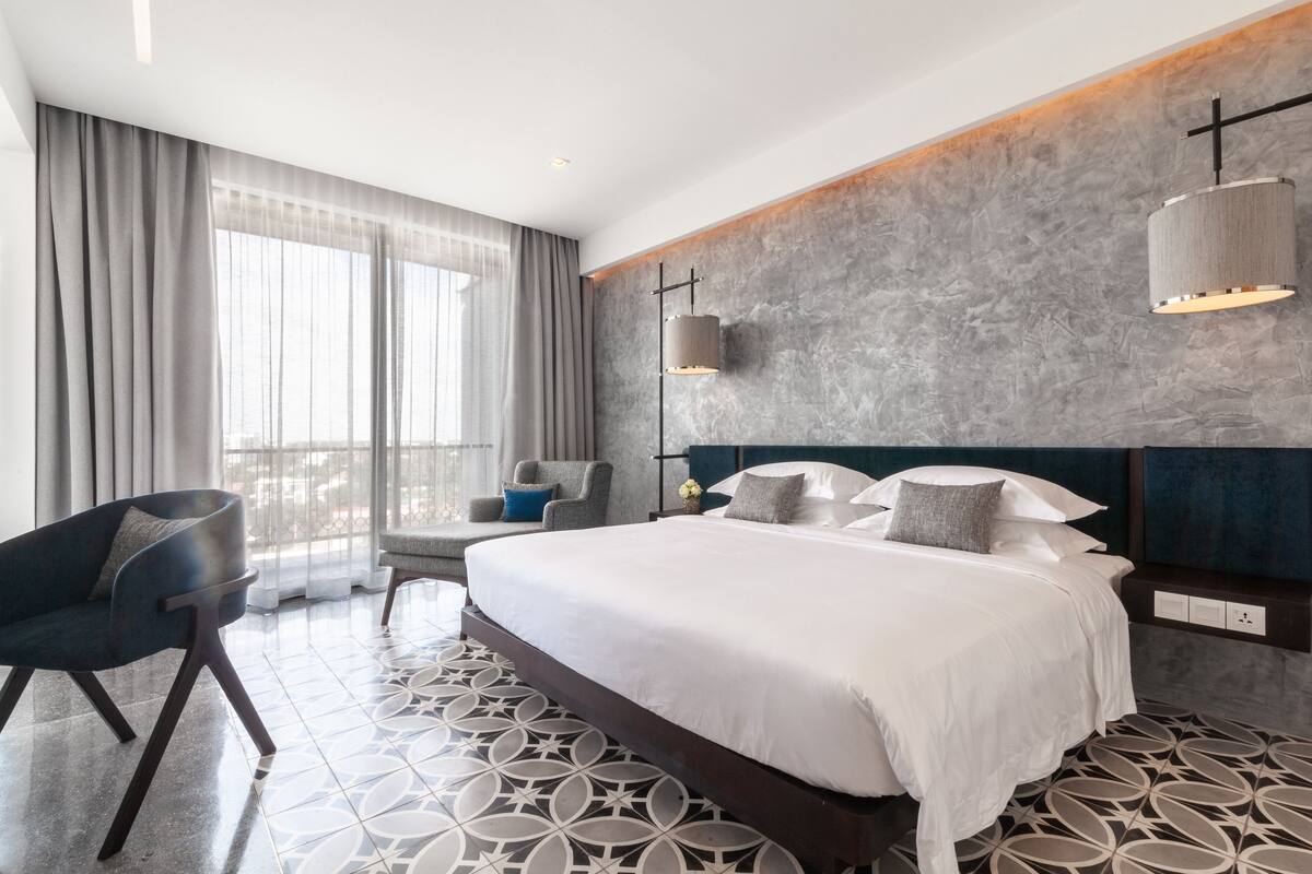 Quarto de hotel moderno com uma cama grande, uma poltrona, uma escrivaninha e vista da cidade através de amplas janelas.