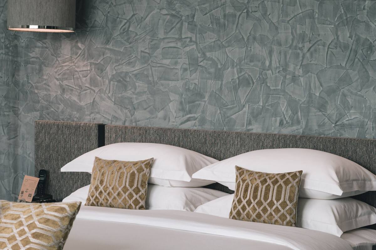 Quarto de hotel moderno com uma cama grande, uma poltrona estofada cinza e uma parede texturizada.