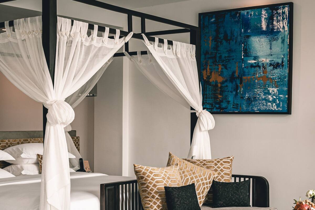 Quarto moderno com cama de dossel, sofá-cama, almofadas estampadas e uma grande pintura abstrata na parede.