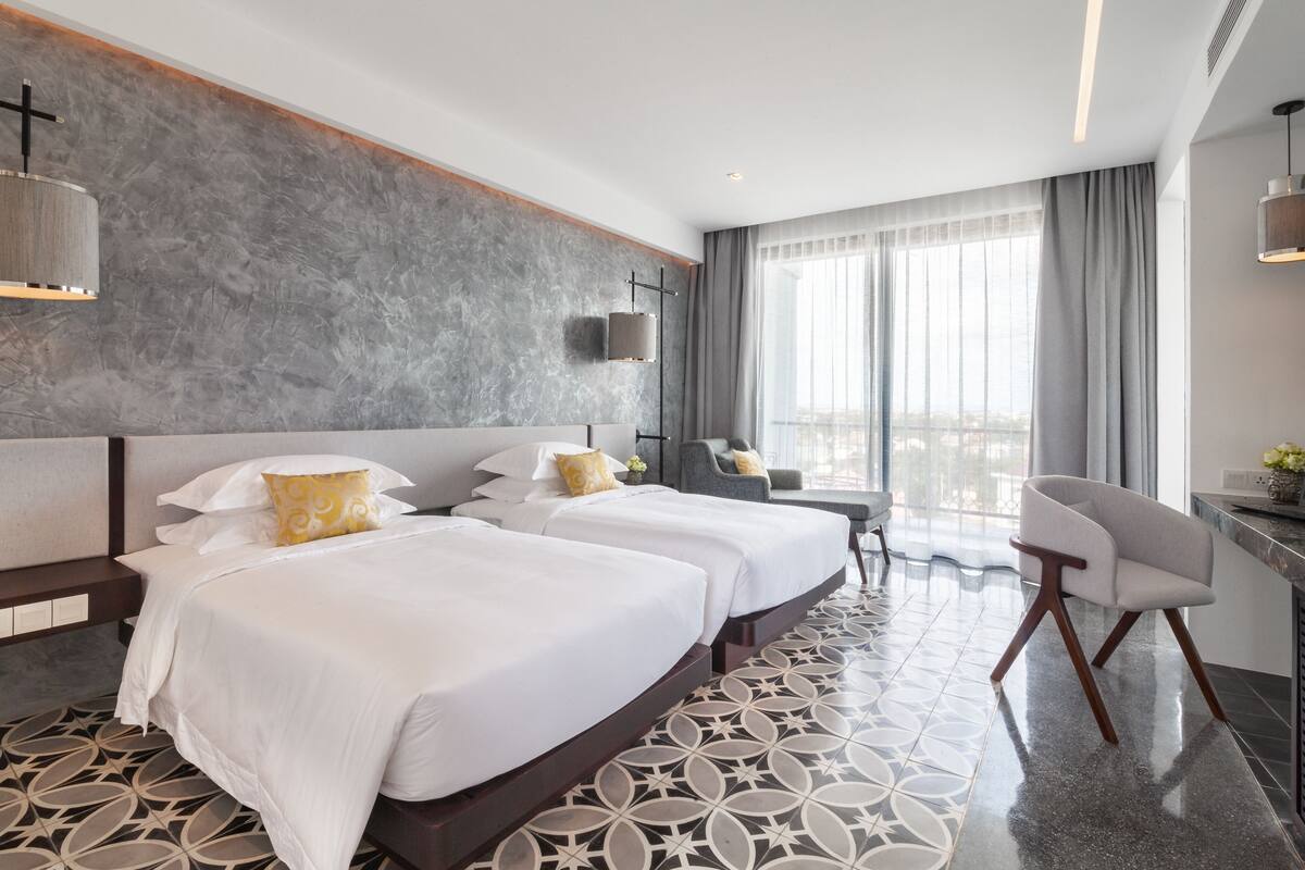 Quarto de hotel com duas camas, uma escrivaninha, uma cadeira e uma janela grande com cortinas.
