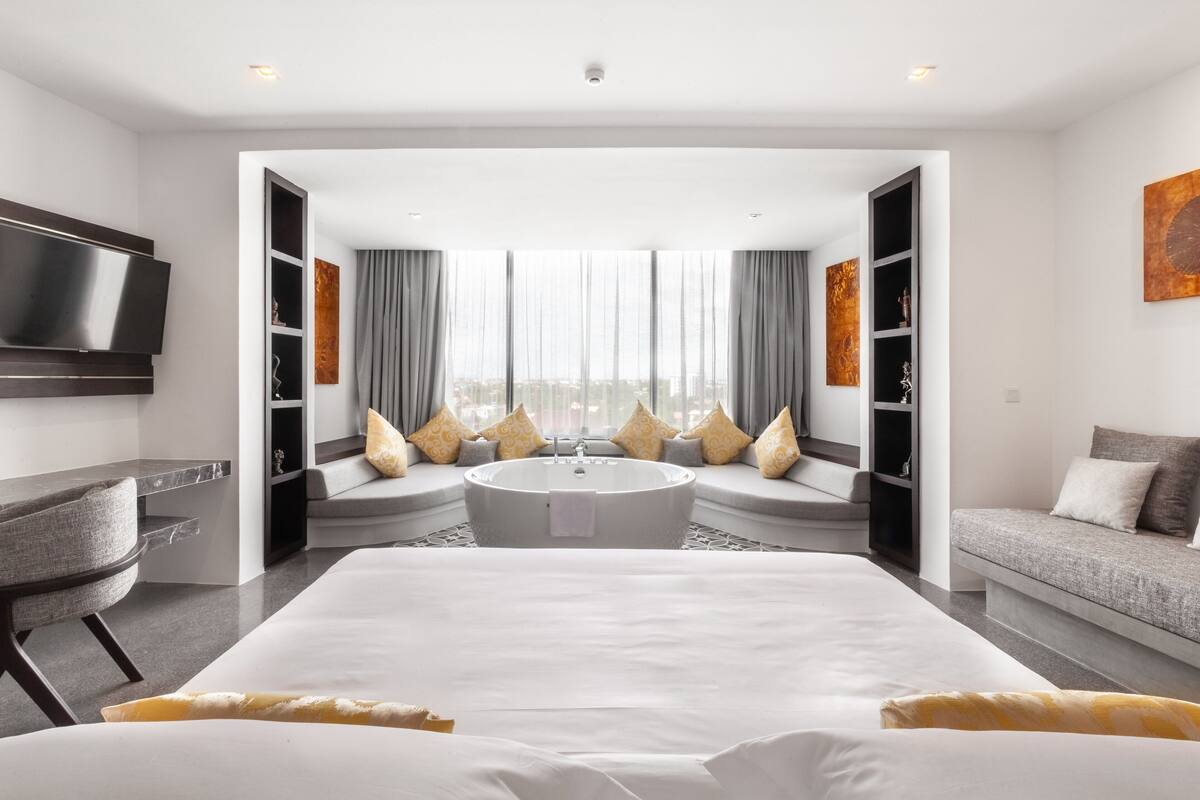 Quarto de hotel moderno com cama grande, banheira redonda, sofá e televisão.