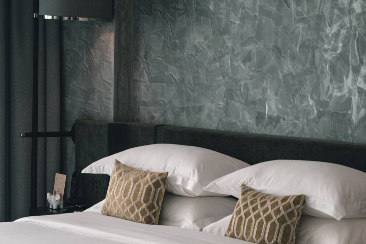 Cama de casal com lençóis brancos e almofadas estampadas, mesa de cabeceira com abajur e parede com textura.