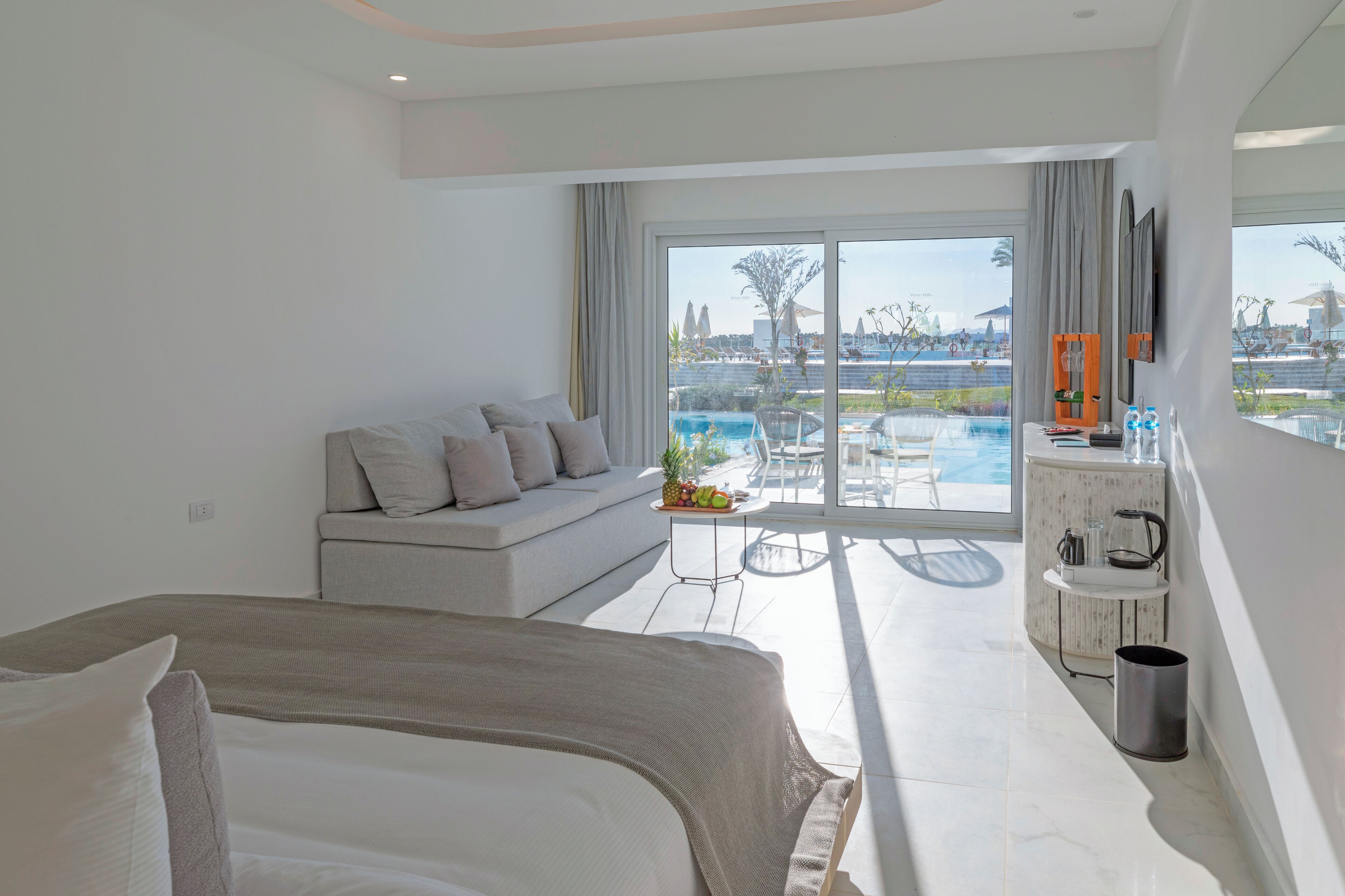 Quarto de hotel com cama, sofá, mesa pequena com frutas, cafeteira e vista para a piscina e para a paisagem urbana.