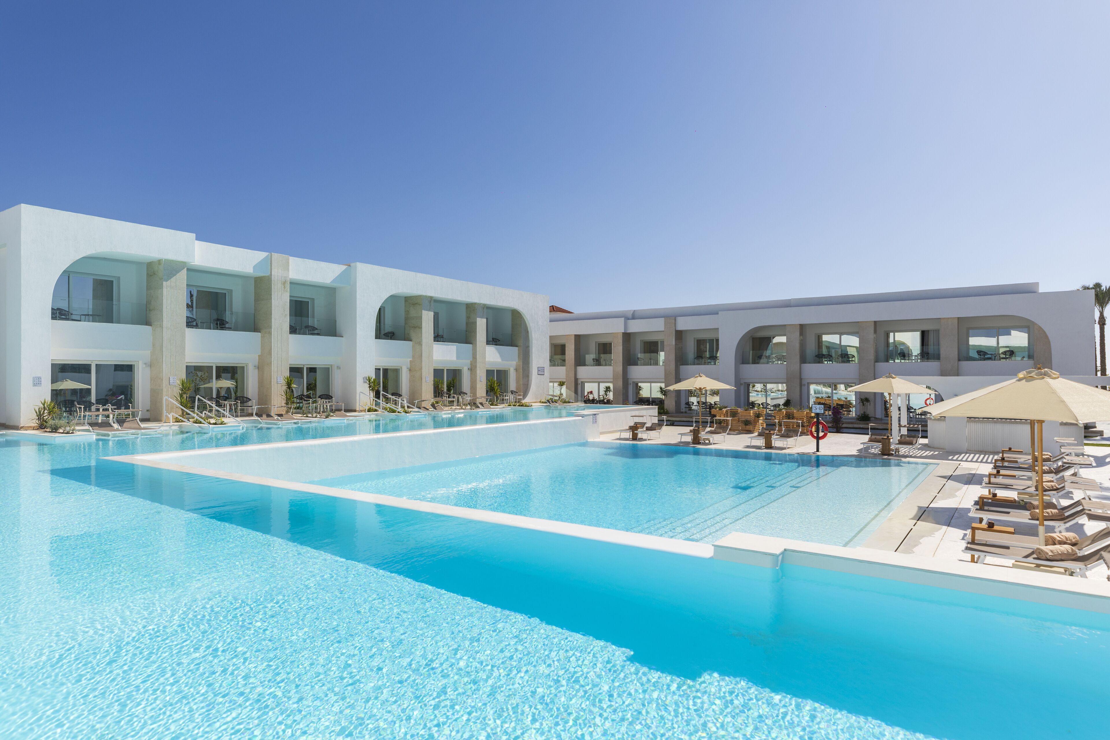 Um hotel moderno com uma piscina grande, várias áreas de estar e um céu azul límpido.