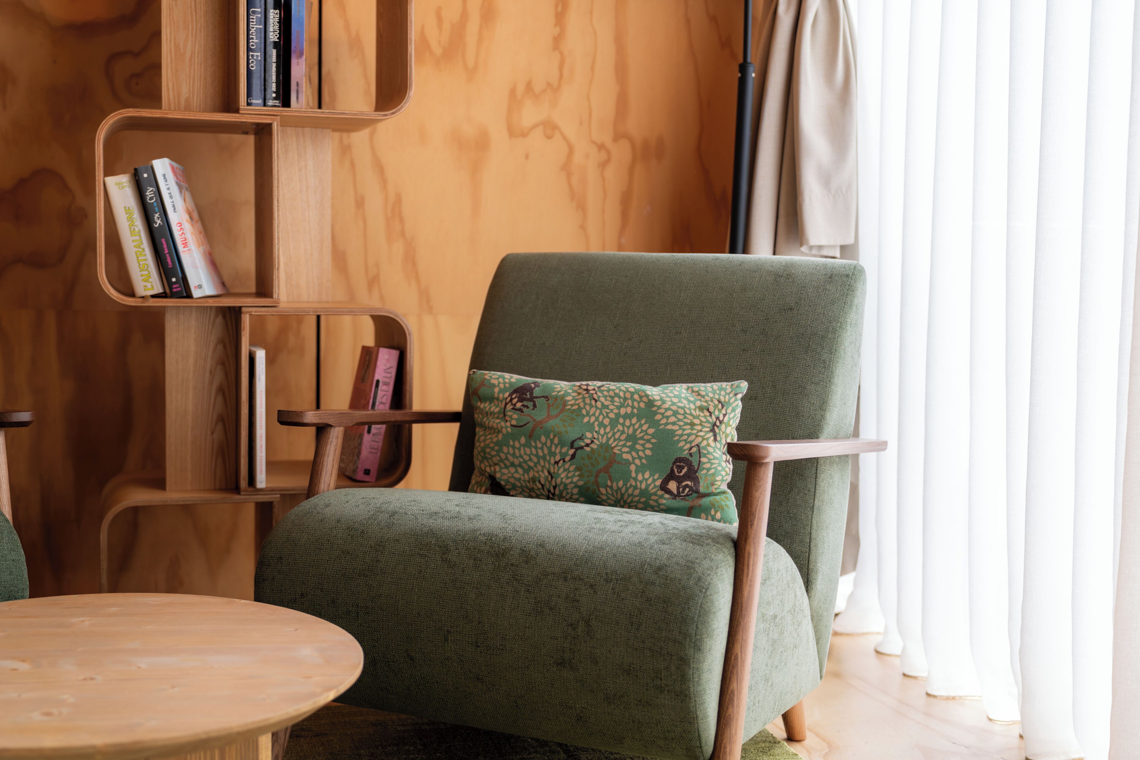 Uma poltrona verde com uma almofada estampada, uma estante de madeira com livros e uma mesa redonda de madeira.