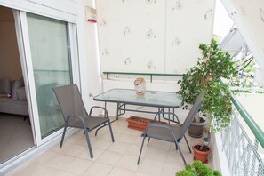 Uma varanda com mesa de vidro, duas cadeiras, um vaso com planta e uma porta de correr de vidro que dá para o interior.