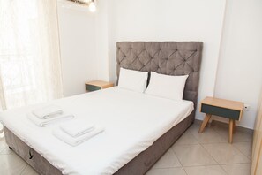 Cama bem arrumada com lençóis brancos e duas almofadas, mesa de cabeceira de madeira com gaveta e luminária fixada na parede.