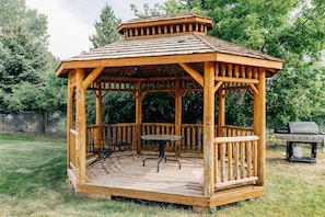 Um gazebo de madeira com uma mesa e cadeiras, cercado por grama e árvores.