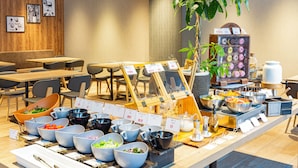 Disposição de buffet com diversas opções de comida em recipientes e utensílios de servir, uma máquina de café e um quadro de cardápio.