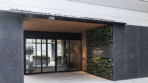 A entrada do Hotel Keihan, com design moderno, apresenta uma porta de vidro, interior de madeira e uma parede verde com plantas.