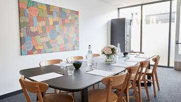 Uma sala de conferências com uma mesa comprida, cadeiras de madeira, uma grande pintura abstrata, um vaso com flores e um armário.