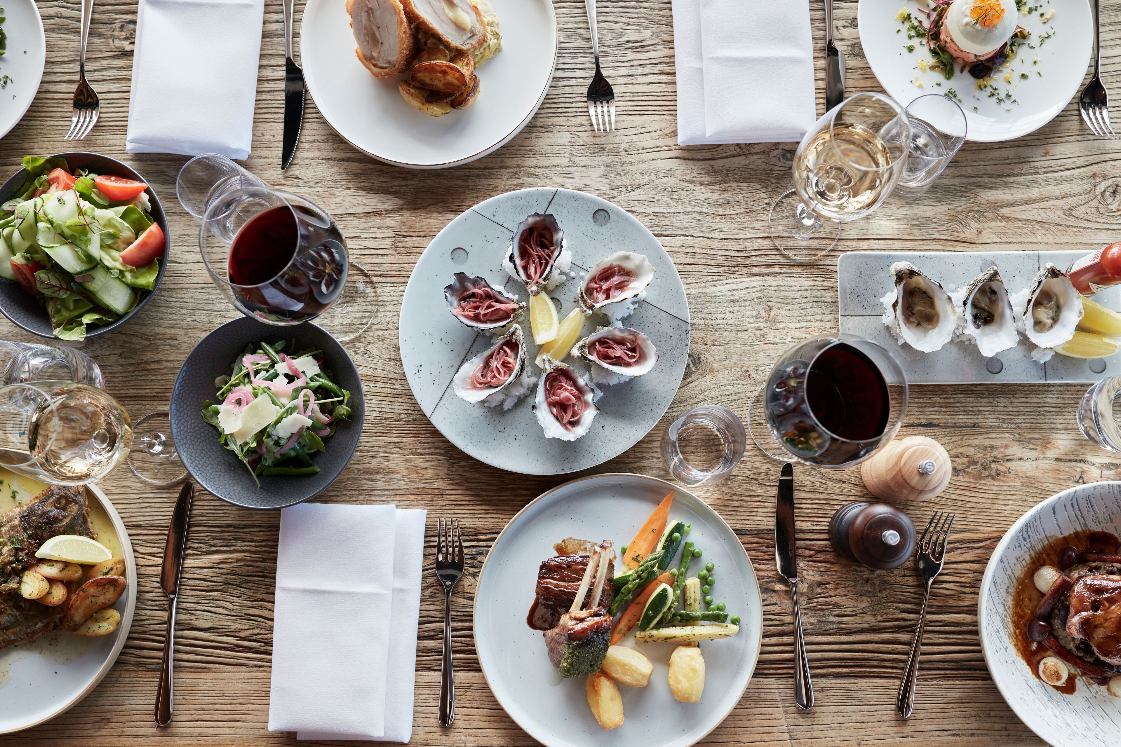 Uma mesa de madeira posta com diversos pratos, incluindo frutos do mar, legumes e carne, acompanhada de copos de vinho e talheres.