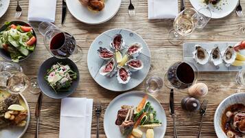 Uma mesa de madeira posta com diversos pratos, incluindo frutos do mar, legumes e carne, acompanhada de copos de vinho e talheres.