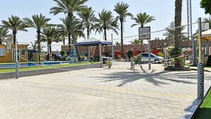 Espaço ao ar livre pavimentado, com palmeiras, quadra de basquete e um prédio com toldo azul.