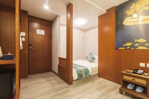 Quarto de hotel com guarda-roupa de madeira, cama com colcha azul e branca, mesa de cabeceira com telefone e abajur, e pintura de peixes pendurada na parede.