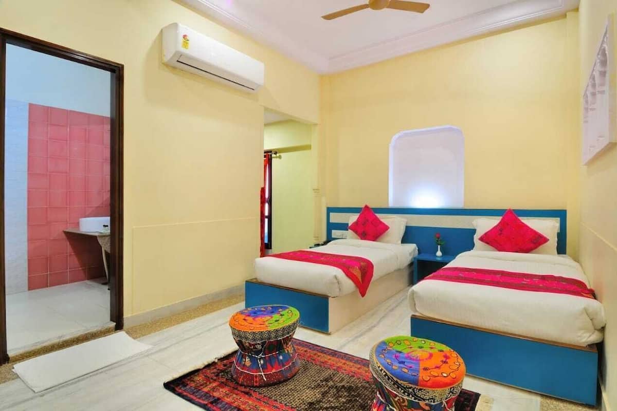 Quarto de hotel com duas camas, um ventilador de teto, aparelho de ar condicionado e um pequeno banheiro visível através de uma porta de vidro.