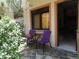 Um pátio externo com mesa e duas cadeiras roxas, janela com cortinas e chão de pedra.