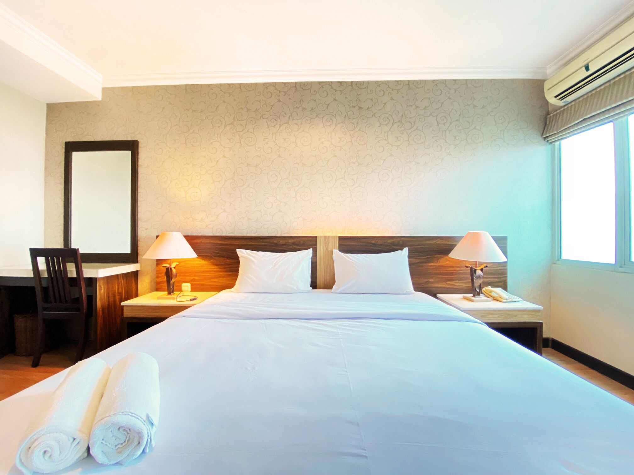 Quarto de hotel com uma cama grande, duas mesas de cabeceira com luminárias, uma cadeira, uma escrivaninha e um espelho grande na parede.