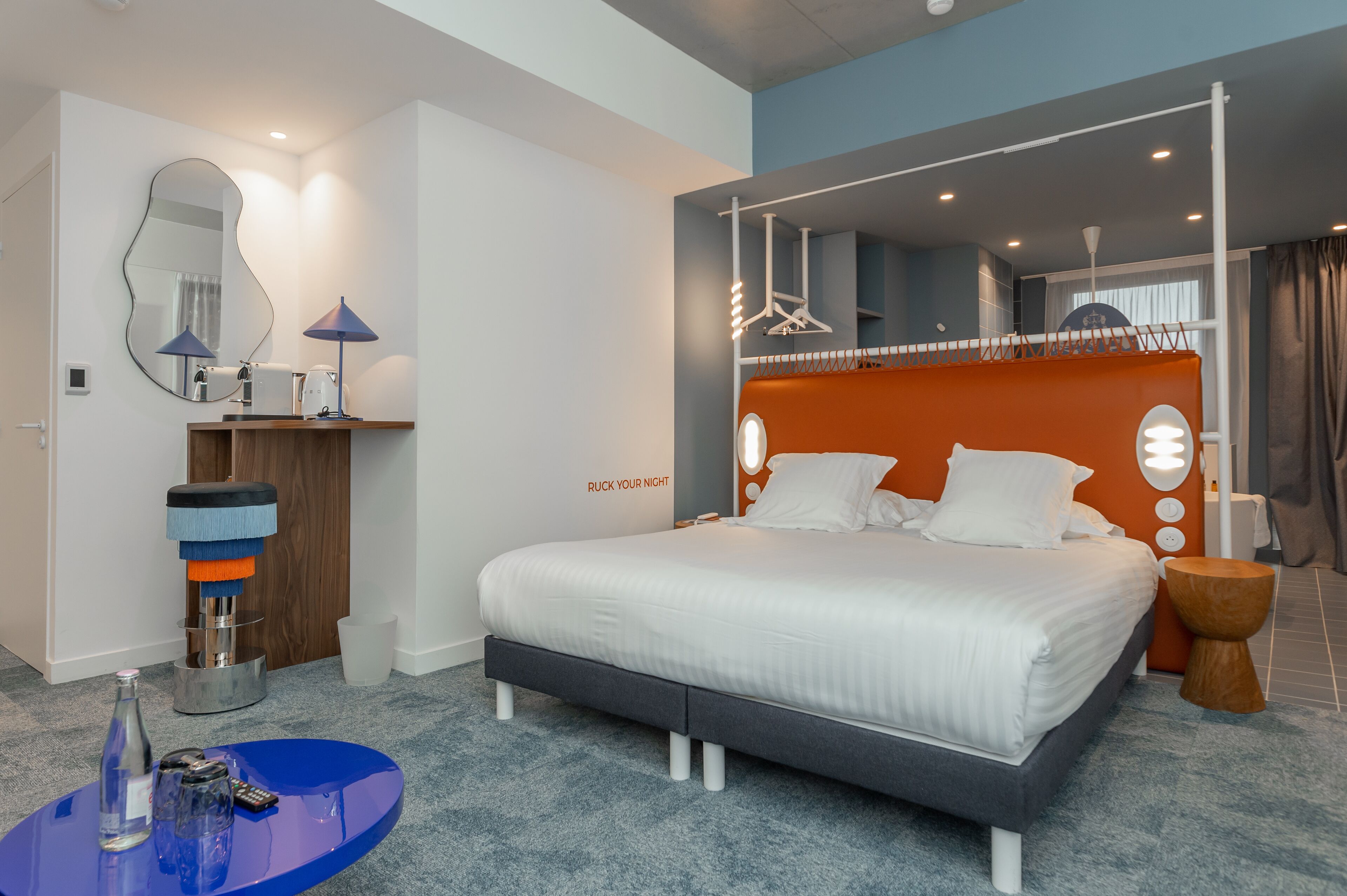 Quarto de hotel moderno com cama grande, mesa de cabeceira, espelho e um espaço pequeno para o banheiro.