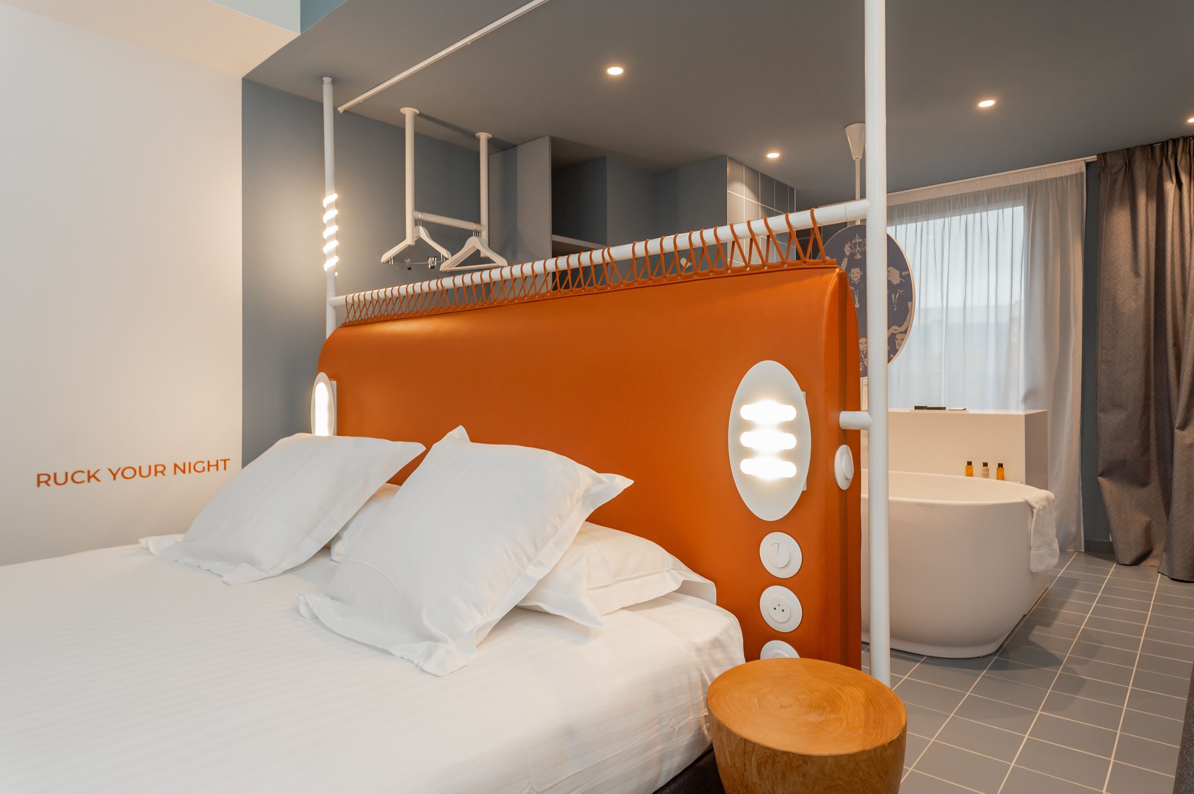 Quarto de hotel moderno com uma cama, um banquinho de madeira, um espelho grande e uma banheira.