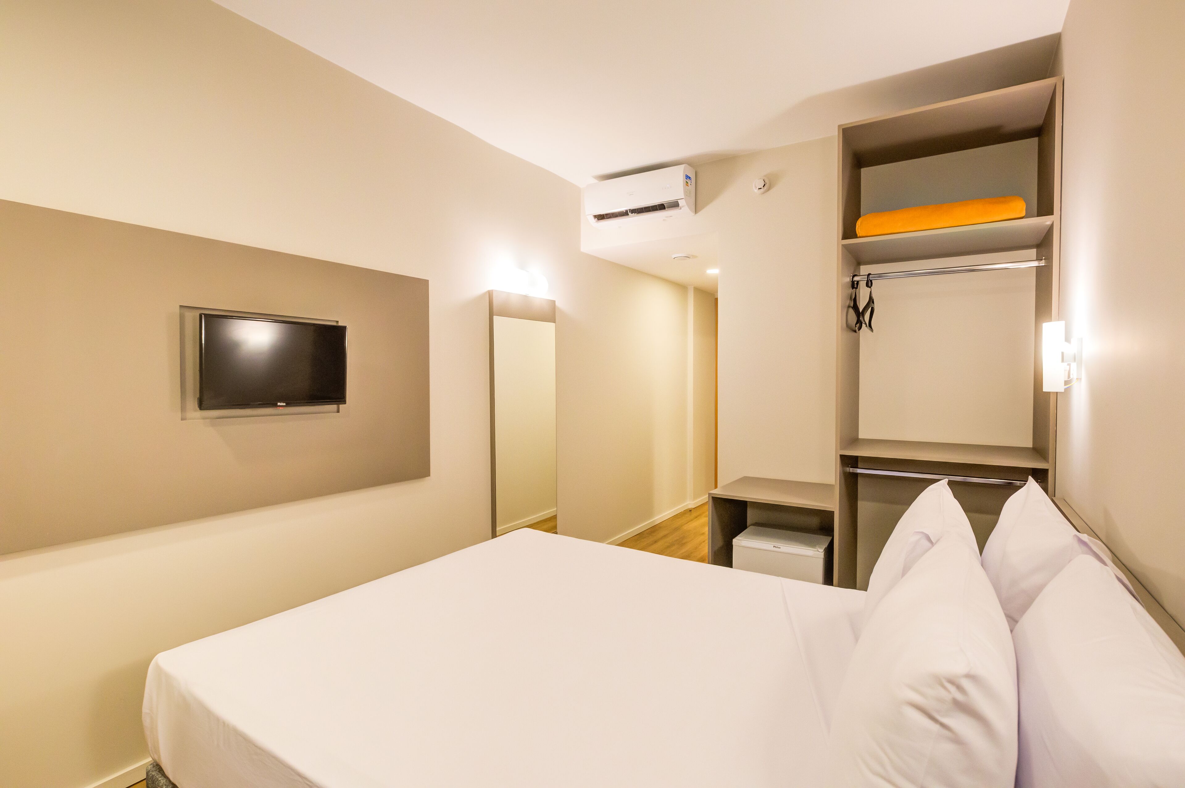 Quarto de hotel com uma cama, televisão fixada na parede, ar condicionado, espelho e um guarda-roupa com prateleiras.