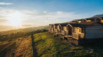Fileira de cabanas de madeira com varandas, localizadas numa encosta gramada com vista para colinas ondulantes ao fundo.